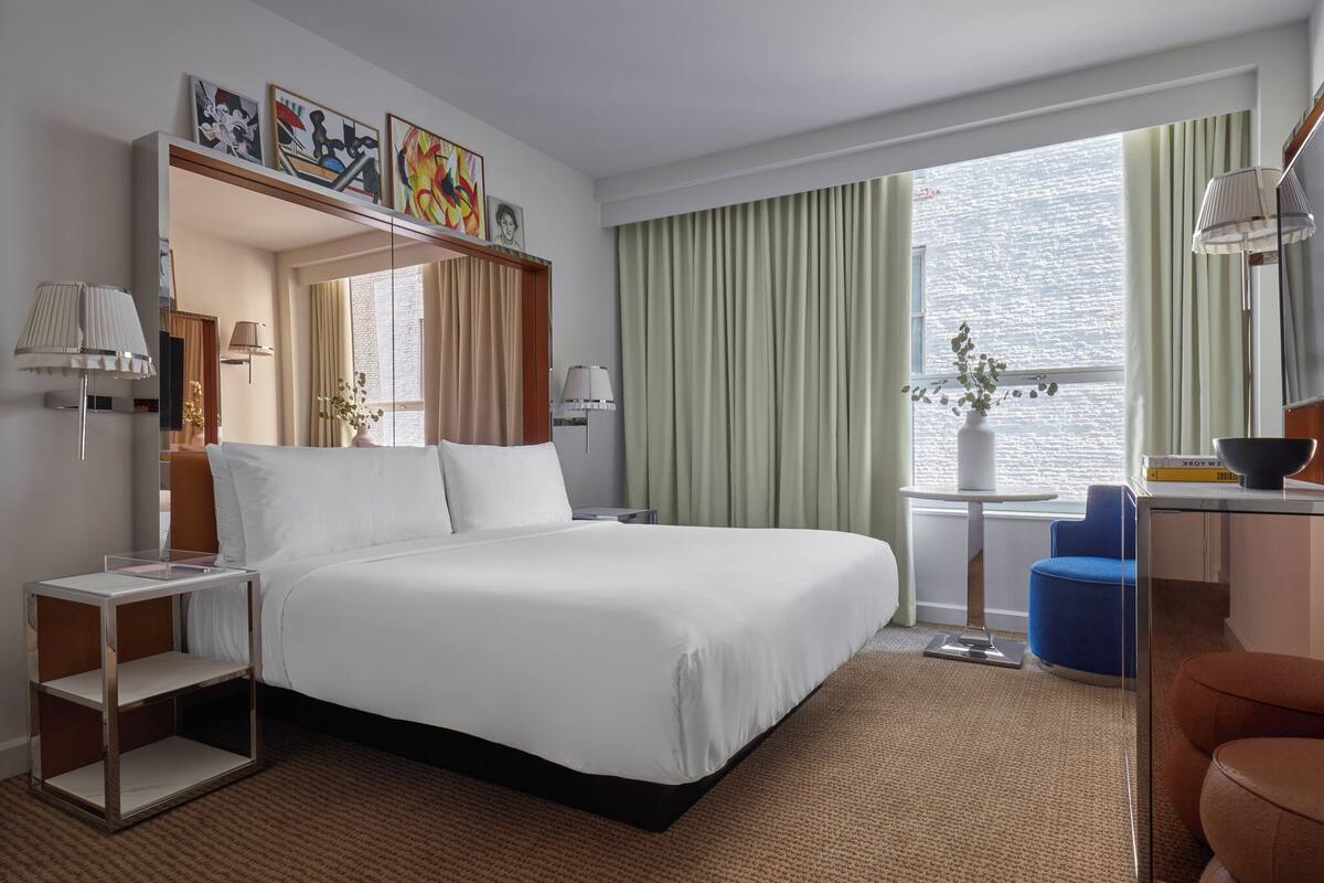 Quarto de hotel com uma cama grande, uma escrivaninha com luminária, uma cadeira e vista para o exterior.