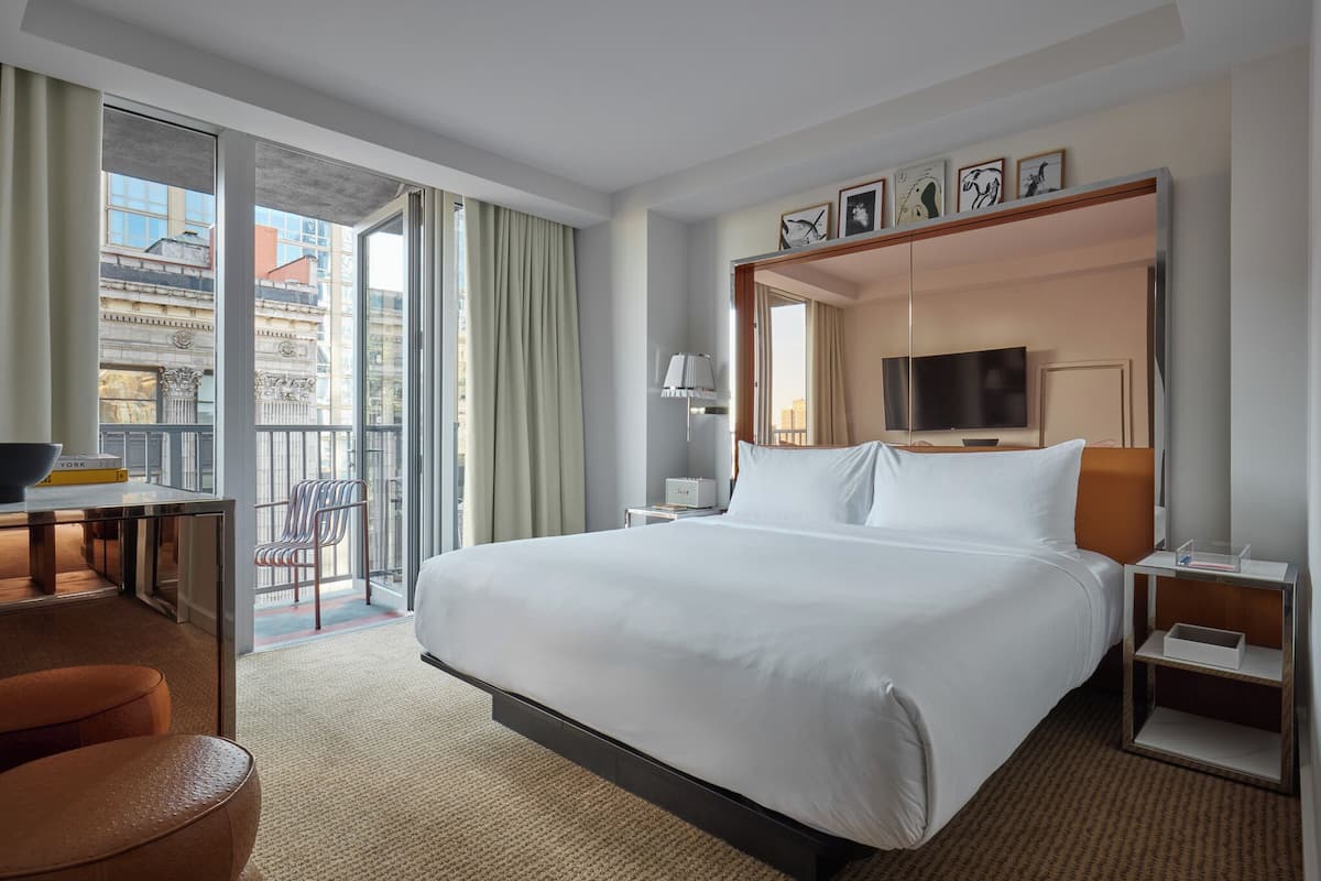 Quarto de hotel com uma cama grande, uma escrivaninha com cadeira, televisão e varanda com vista para edifícios.