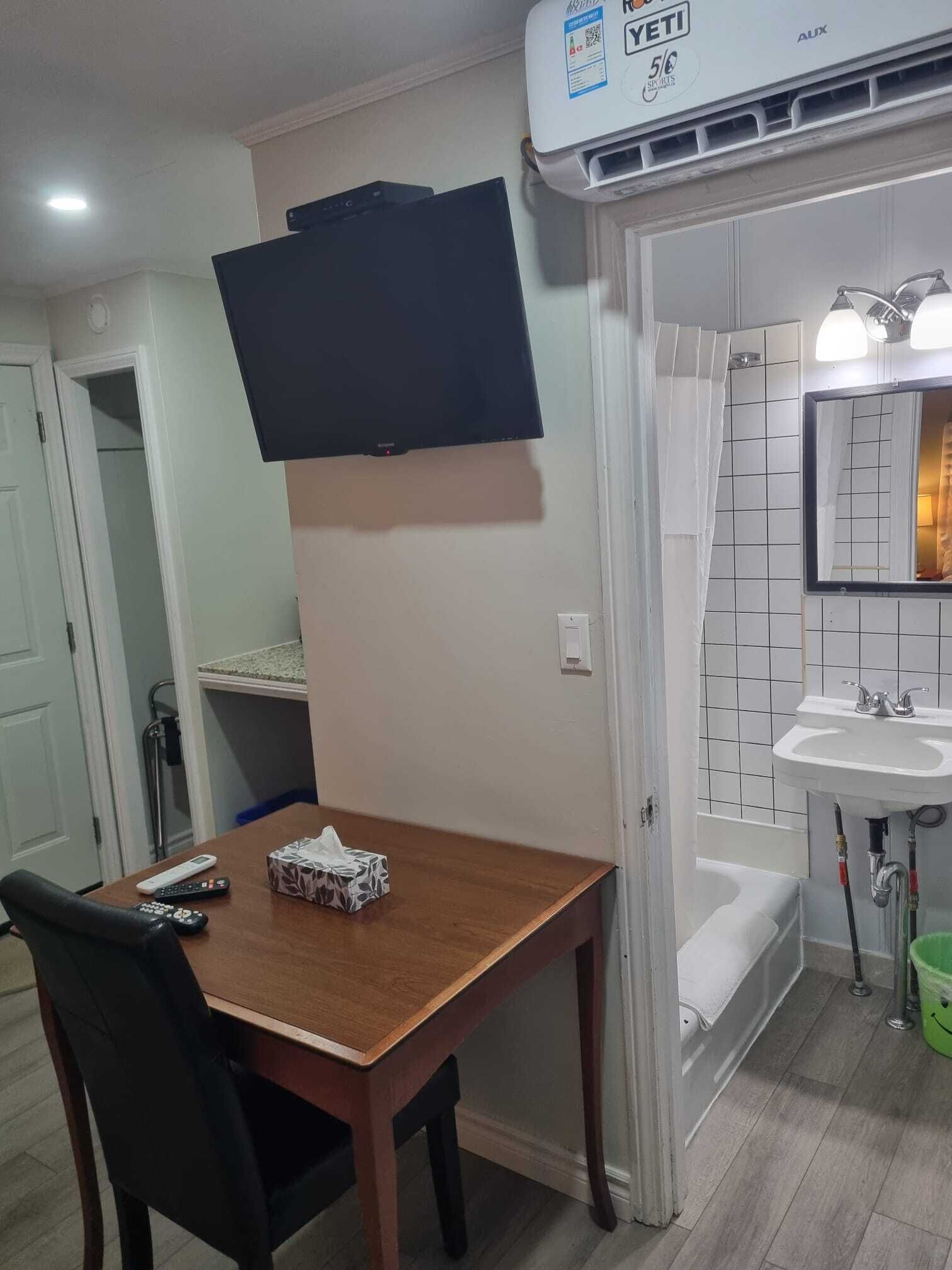 Quarto com televisão presa na parede, uma mesa com uma caixa e controles remotos, uma cadeira, uma banheira e uma pia.
