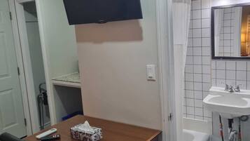 Quarto com televisão presa na parede, uma mesa com uma caixa e controles remotos, uma cadeira, uma banheira e uma pia.