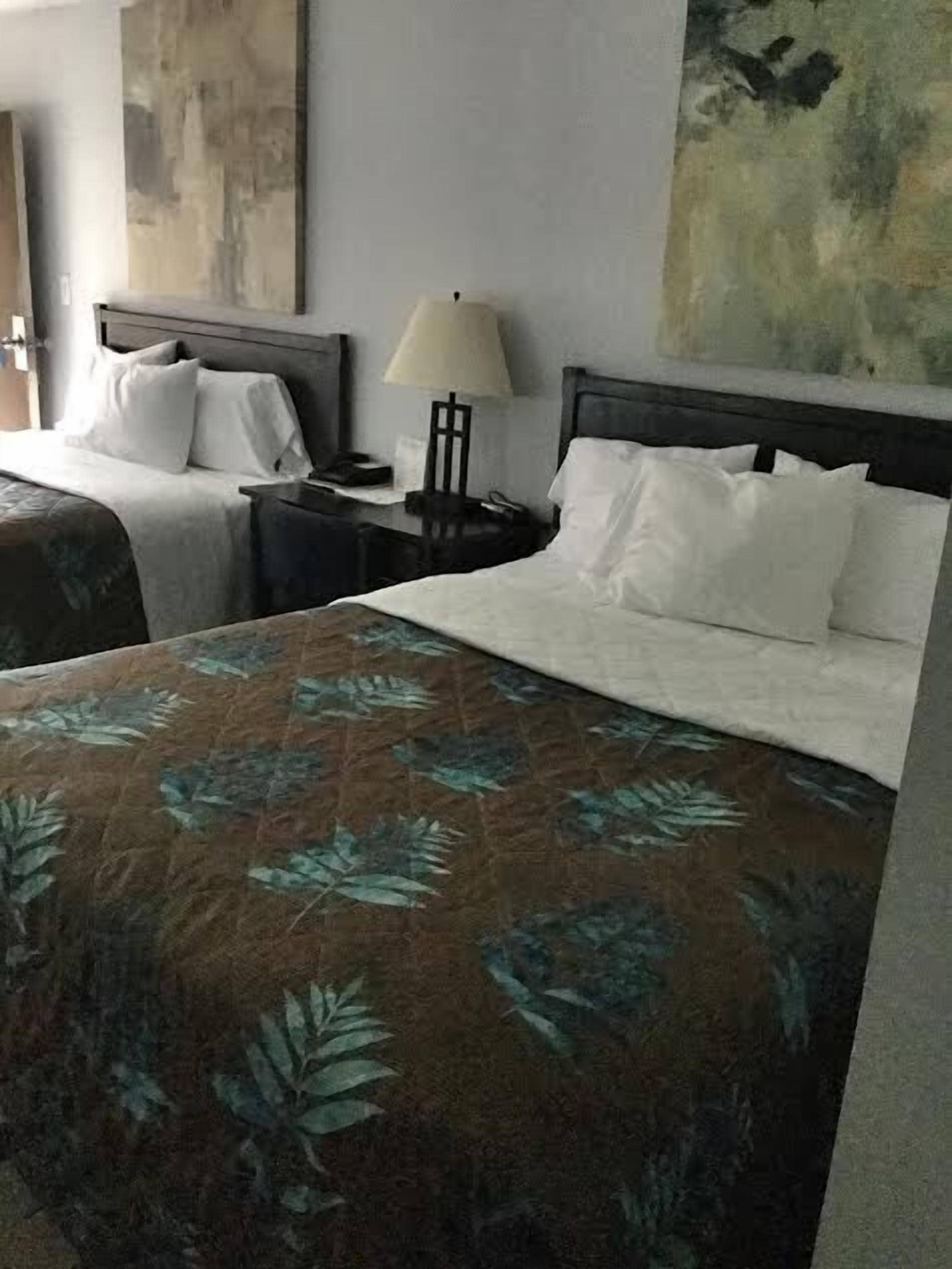 Quarto de hotel com duas camas, uma mesa de cabeceira com luminária e uma grande pintura abstrata na parede.