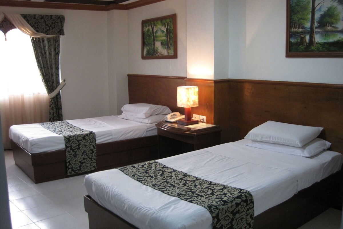 Quarto de hotel com duas camas, mesa de cabeceira com abajur, quadro na parede e janela com cortinas.