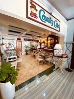 Um café campestre com uma placa convidativa, uma área de jantar com cadeiras e mesa de madeira, e uma cozinha visível ao fundo.