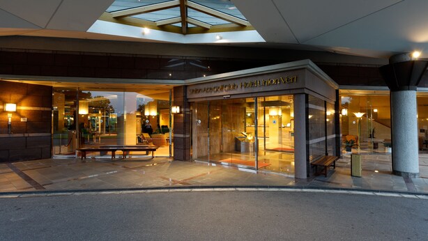 Entrada de hotel com fachada de vidro, um banco do lado de fora e escadas levando à entrada.