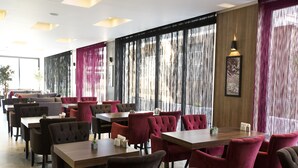Um restaurante com várias mesas e cadeiras, janelas amplas com cortinas e decoração moderna.