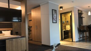 Quarto de hotel moderno com cama, armário de madeira, uma pequena mesa com um vaso decorativo, TV fixada acima da cama e área de kitchenette.