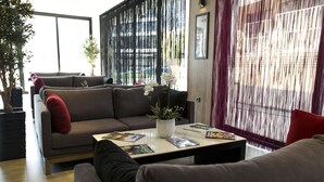 Uma sala de estar moderna com um sofá cinzento, uma mesa de centro com revistas, um vaso de planta e uma janela ampla com cortinas leves.