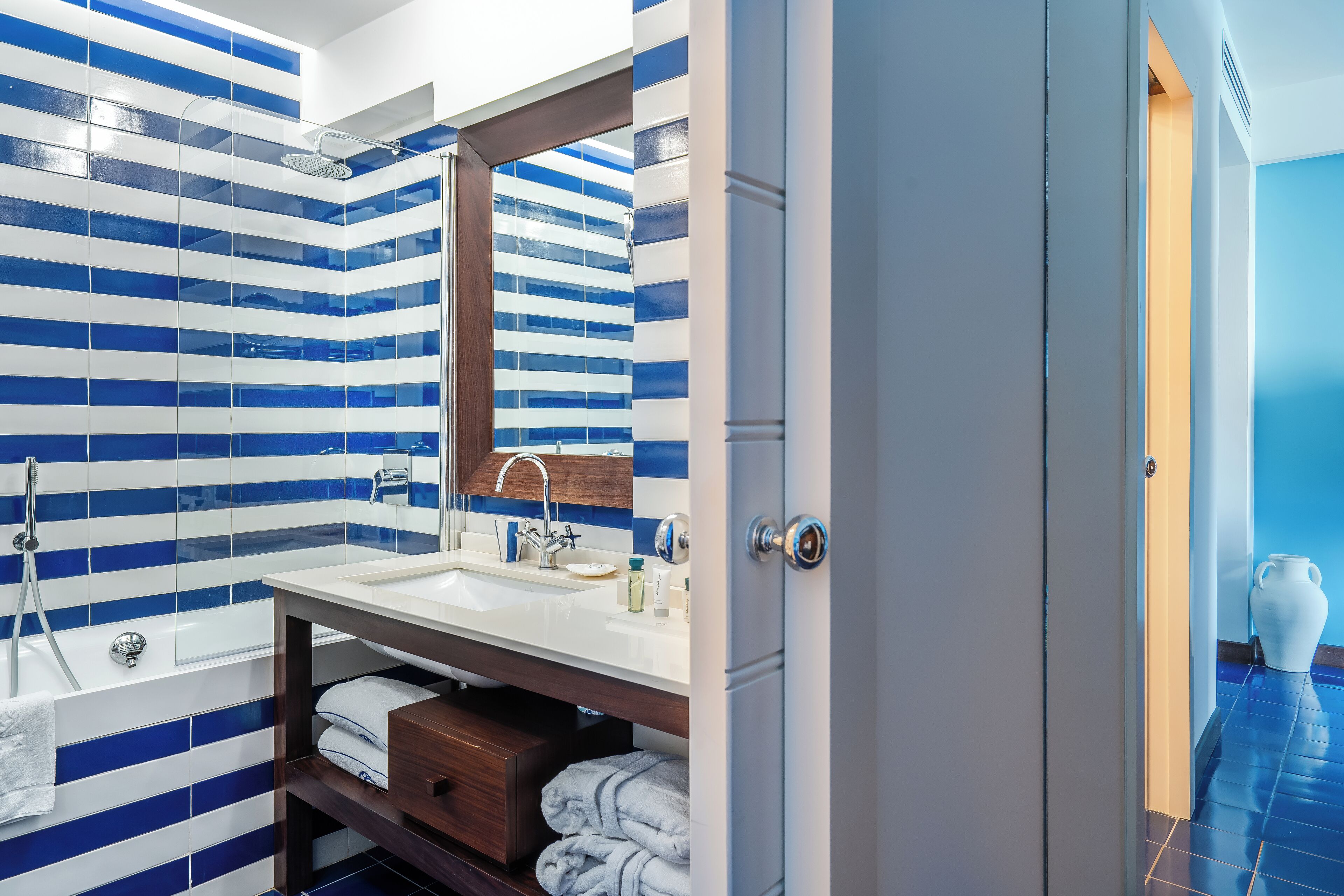 Uma casa de banho moderna com um painel de azulejos listrados em azul e branco, uma pia branca, um espelho e um chuveiro.