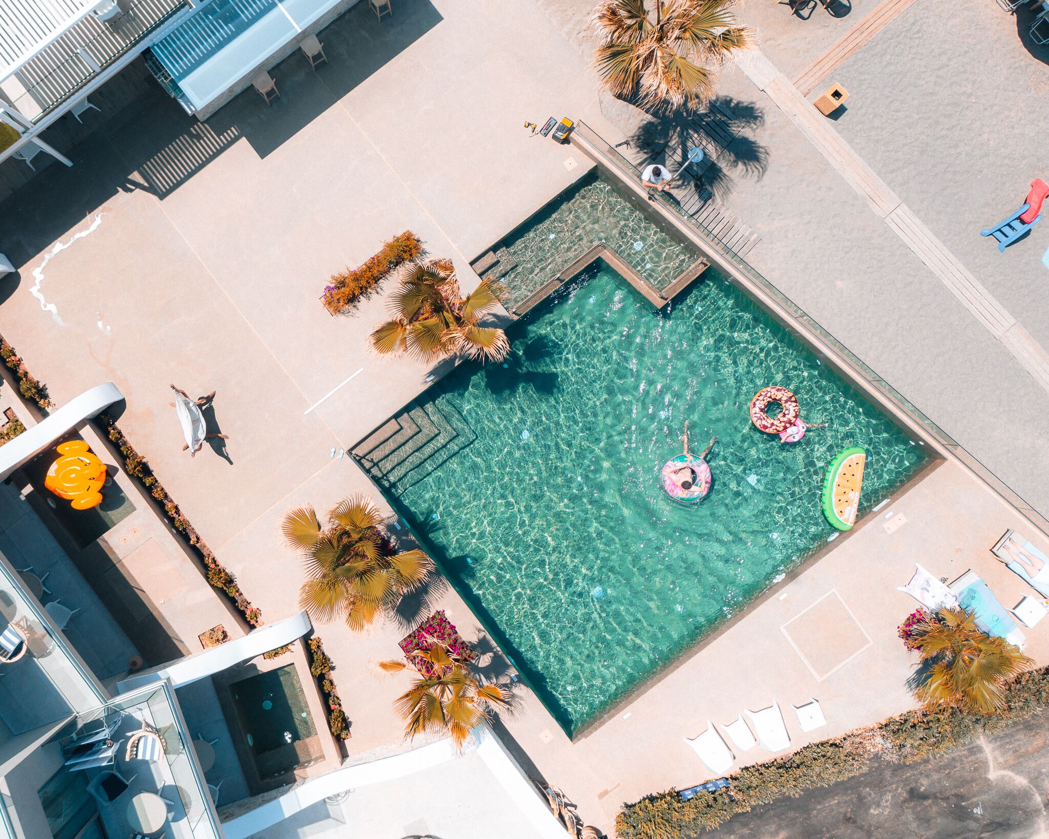 Vista aérea de uma área de piscina com água cristalina, cercada por palmeiras e cadeiras de descanso.