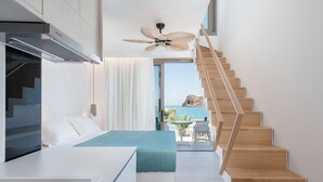 Quarto de hotel moderno com cama, escrivaninha, ventilador de teto, escada e vista para o mar.