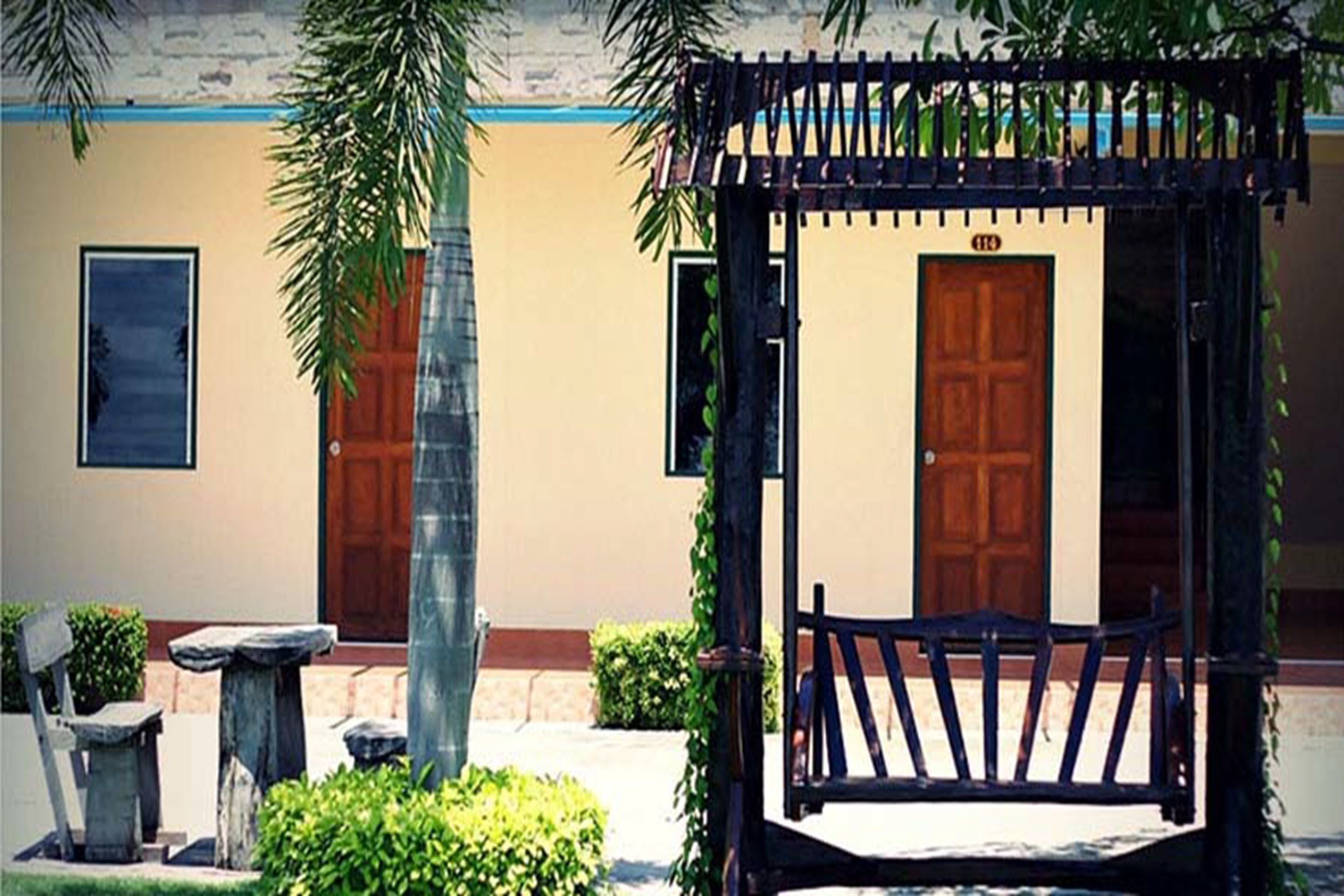Pátio com pérgola de madeira, um banco de pedra e um vaso de pedra. Há duas portas e uma janela no edifício.