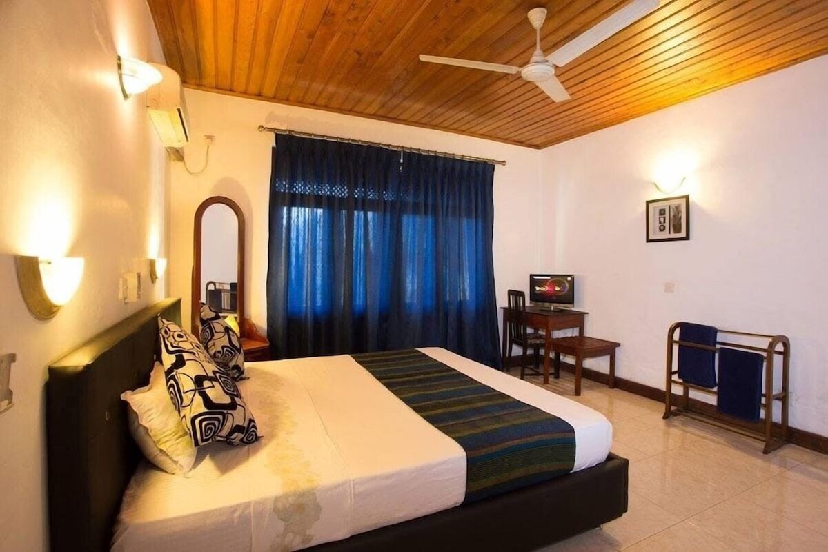 Quarto com cama, escrivaninha, cadeira, ventoinha no teto e janela com cortinas.