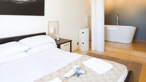 Quarto de hotel com cama, mesa de cabeceira, abajur, toalha, secador de cabelo e banheira.