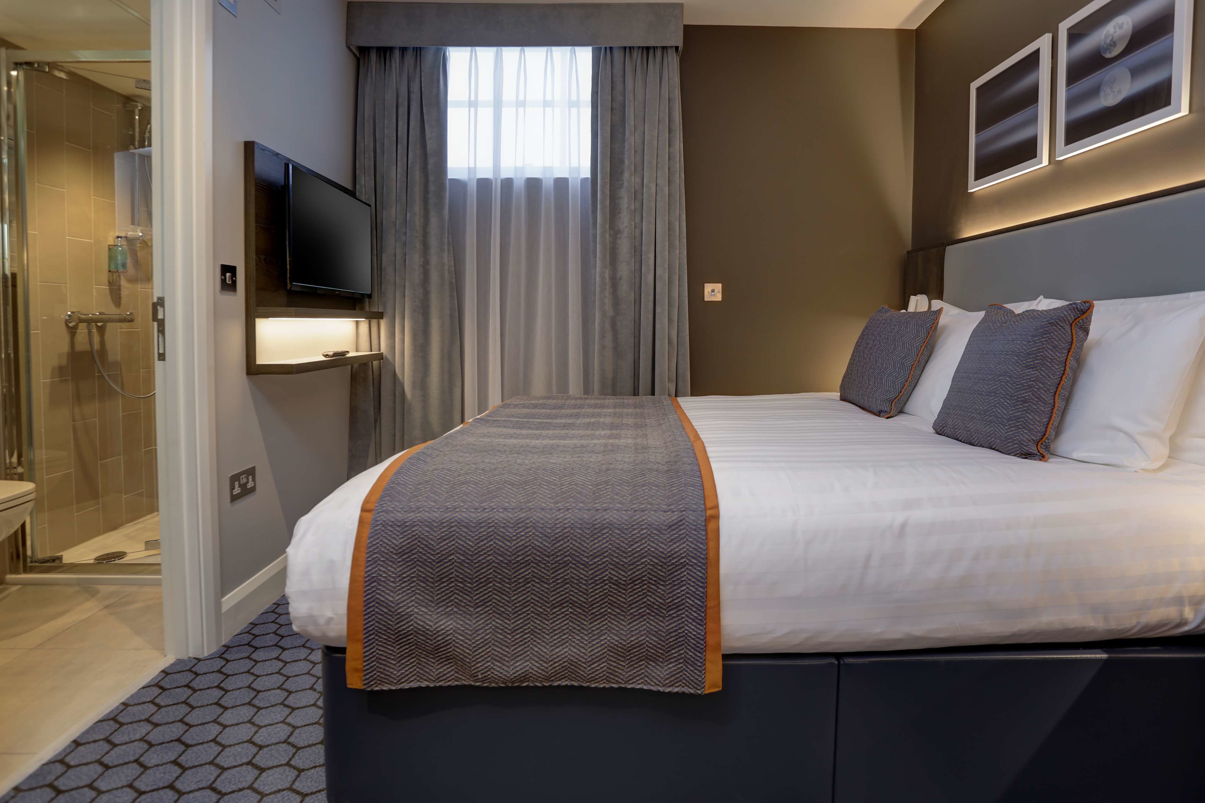 Quarto de hotel moderno com uma cama grande, televisão presa à parede, área de chuveiro e vista para a cidade.
