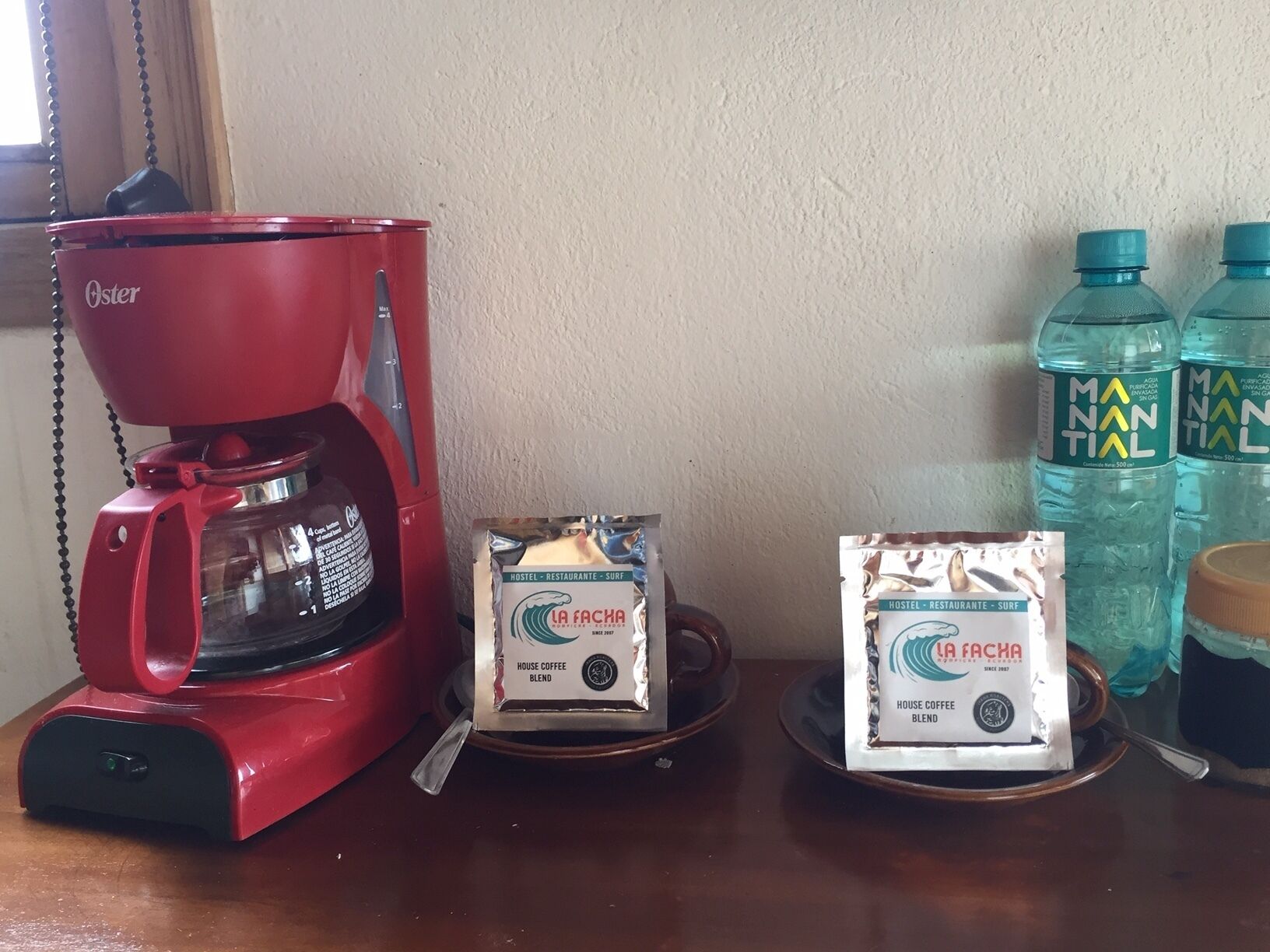 Uma cafeteira vermelha, dois sachês do café da casa La Facha e duas garrafas de água sobre uma mesa.