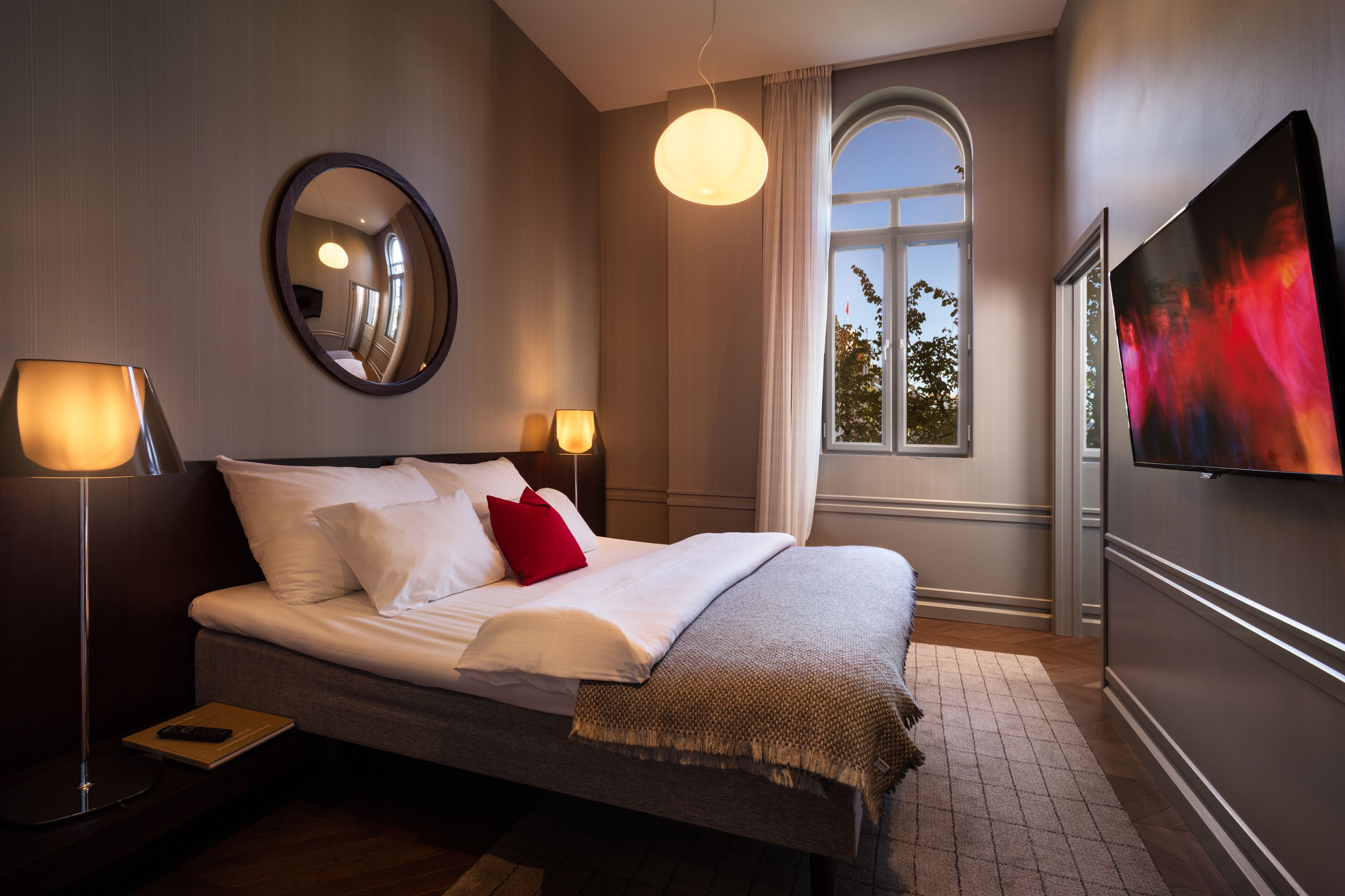 Quarto de hotel moderno com uma cama grande, abajures, um espelho, uma televisão e uma janela com vista para árvores.