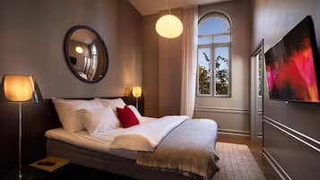 Quarto de hotel moderno com uma cama grande, abajures, um espelho, uma televisão e uma janela com vista para árvores.