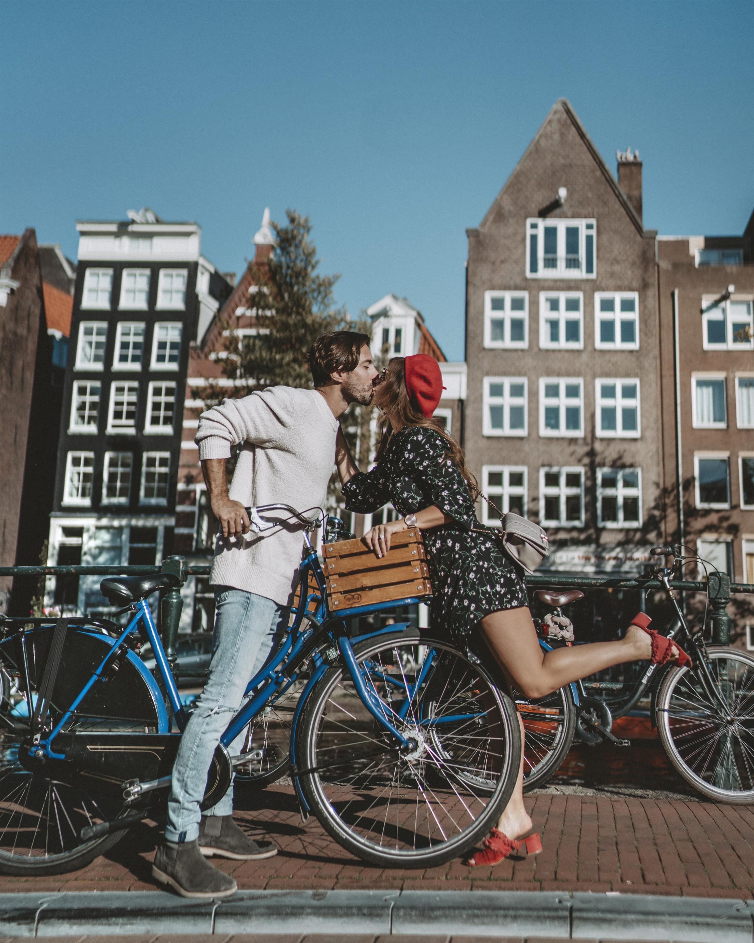 Um casal se beijando em uma bicicleta em frente a uma fileira de edifícios europeus tradicionais.