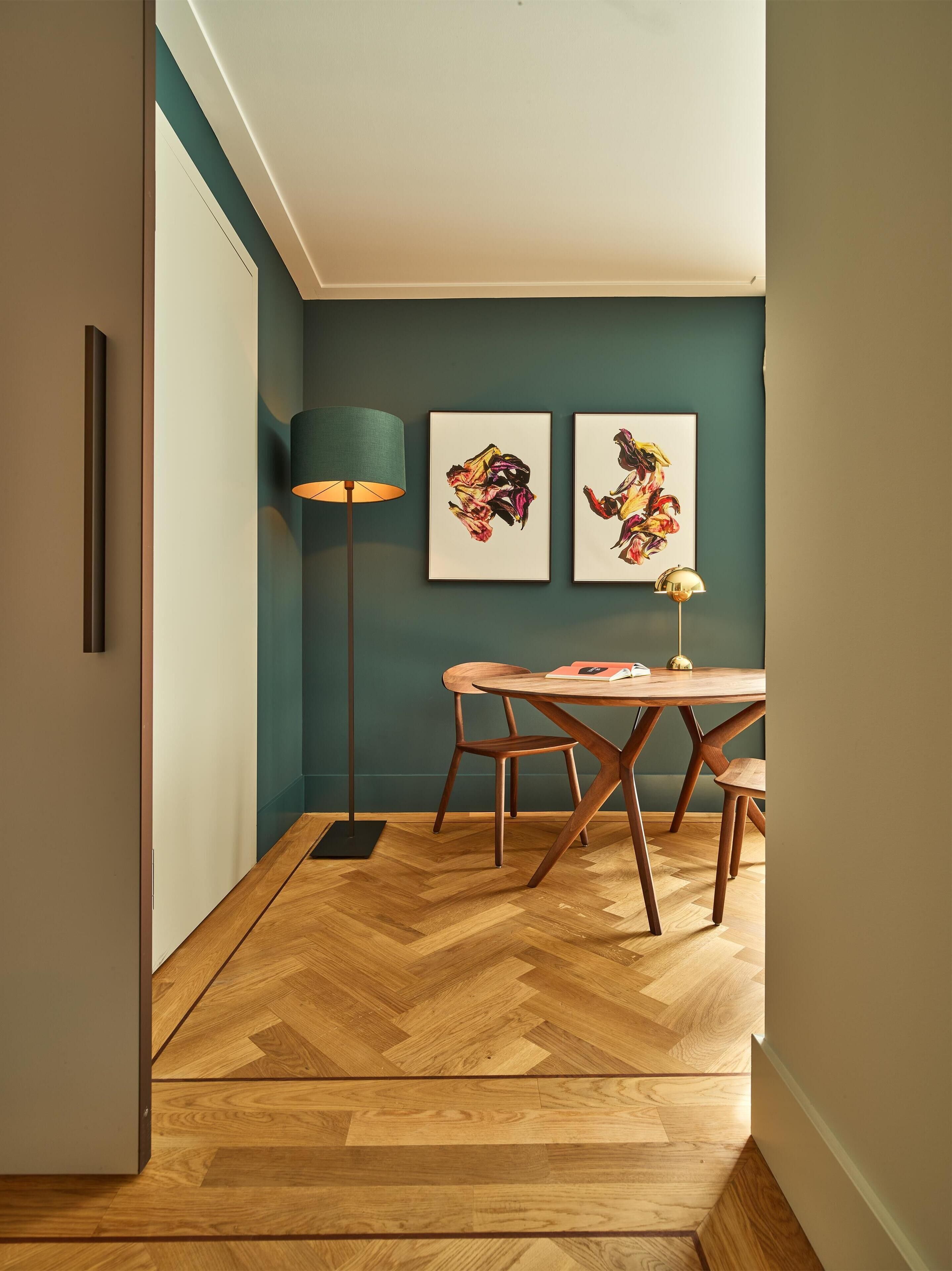 Uma área de refeições com piso de madeira em padrão herringbone, uma mesa redonda e duas cadeiras. Há duas obras de arte penduradas na parede e um abajur.