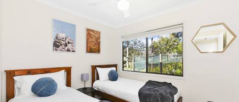 Quarto com duas camas, ventilador de teto, janela com vista para vegetação e obras de arte nas paredes.
