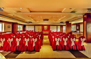 Uma sala de conferências de hotel com fileiras de cadeiras vermelhas com laços brancos, um palco na frente e uma área de recepção à direita.