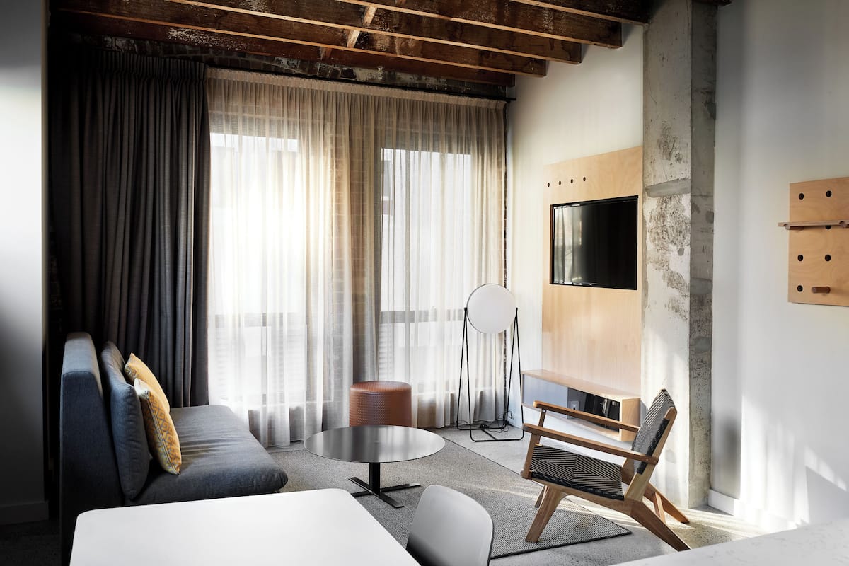 Quarto de hotel moderno com sofá, mesa de centro redonda, cadeira de madeira, um pequeno banquinho e TV fixada na parede.
