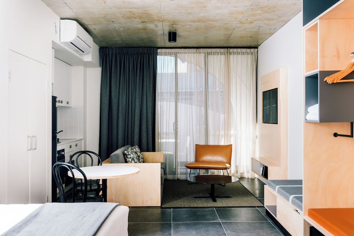 Quarto de hotel moderno com área de jantar, sofá, poltrona, televisão e armário.