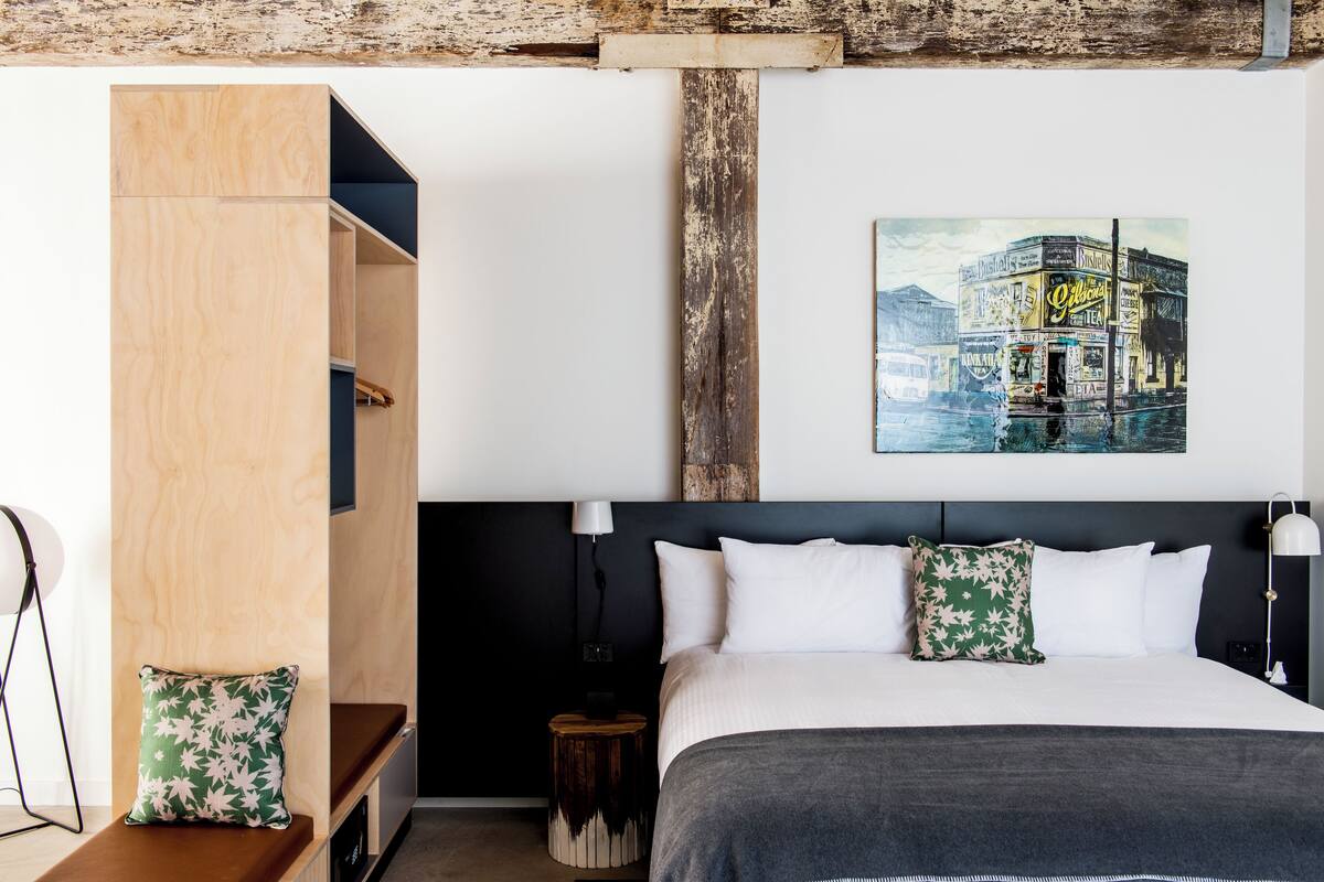 Quarto com cama, guarda-roupa de madeira, um banquinho de madeira e um quadro na parede.