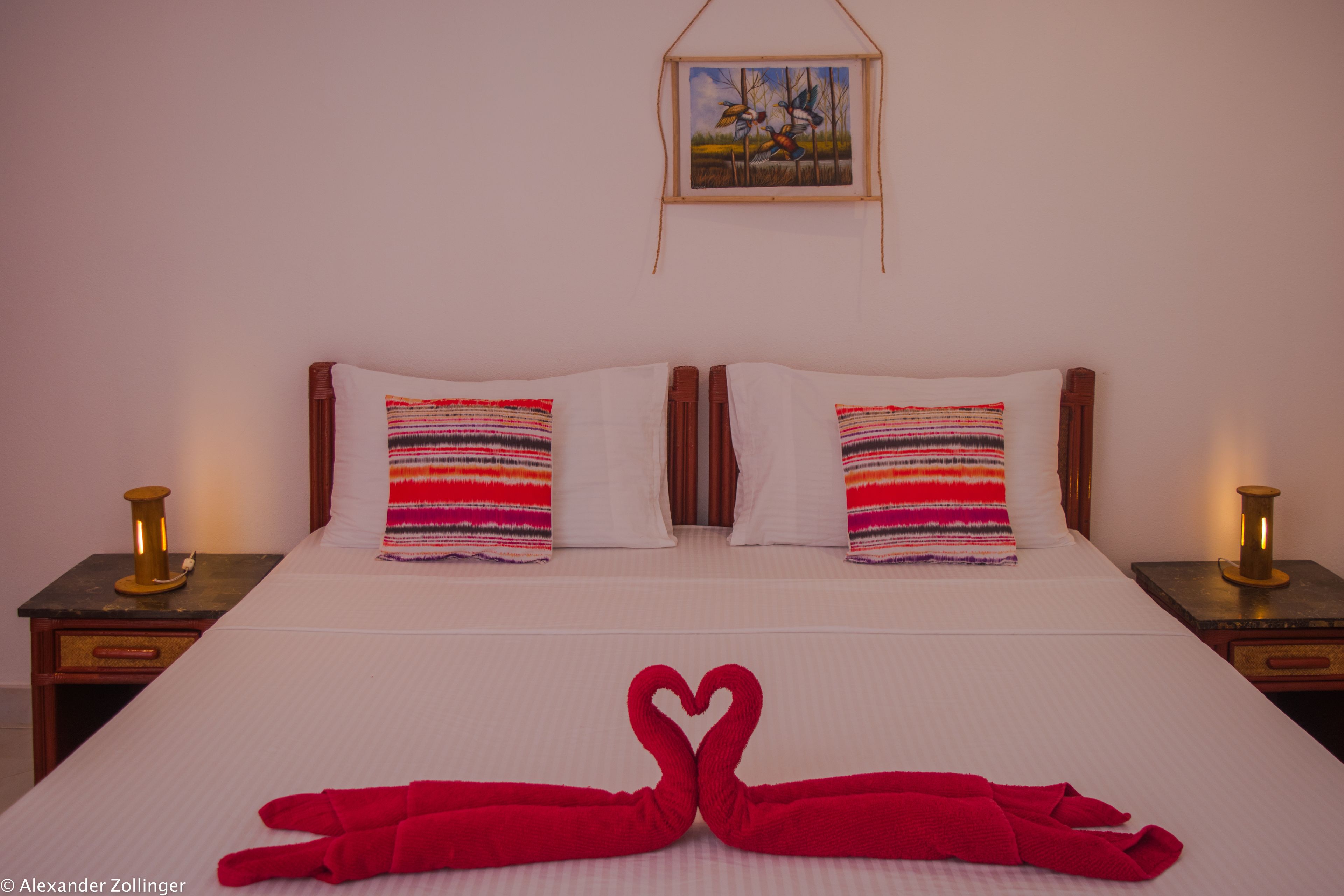 Uma cama bem arrumada com lençóis brancos e duas almofadas, uma listrada e outra lisa, em um quarto com cabeceira e mesas de cabeceira de madeira simples.