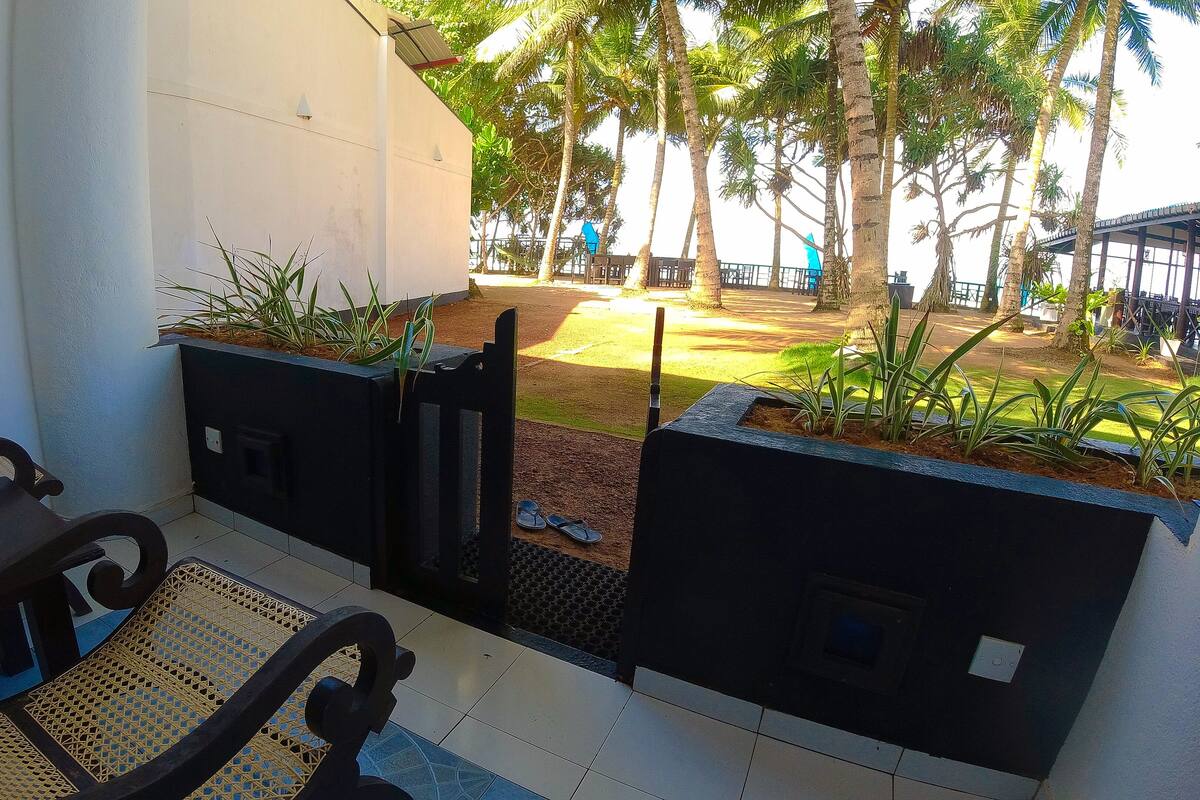 Uma varanda com cadeira e mesa pequena, um portão que dá para um jardim com palmeiras, e um edifício ao fundo.