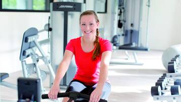 Uma pessoa numa bicicleta ergométrica numa academia, com equipamento de ginástica ao fundo.