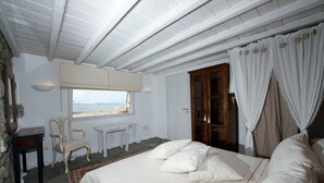 Um quarto com cama, janela com vista, cadeira, mesinha e guarda-roupa.