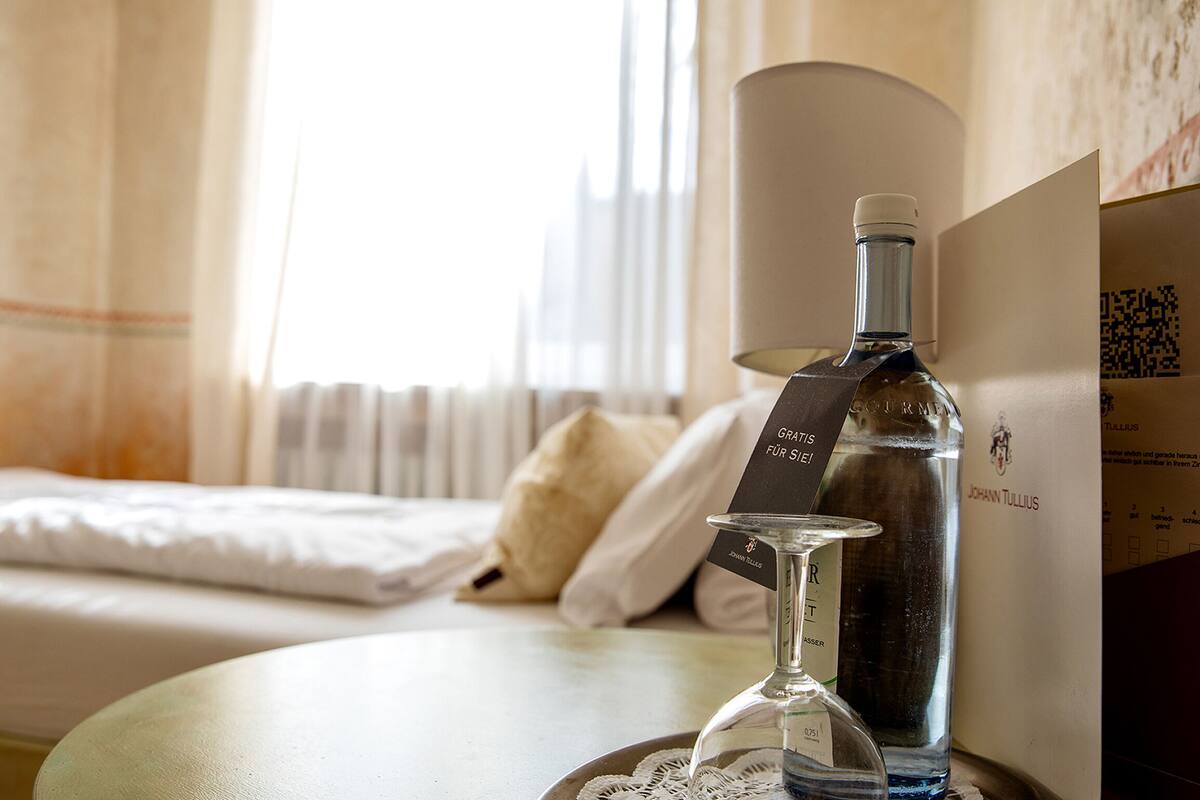 Quarto de hotel com uma cama, uma mesa de cabeceira com uma garrafa e um copo, e um código QR em um cartão.