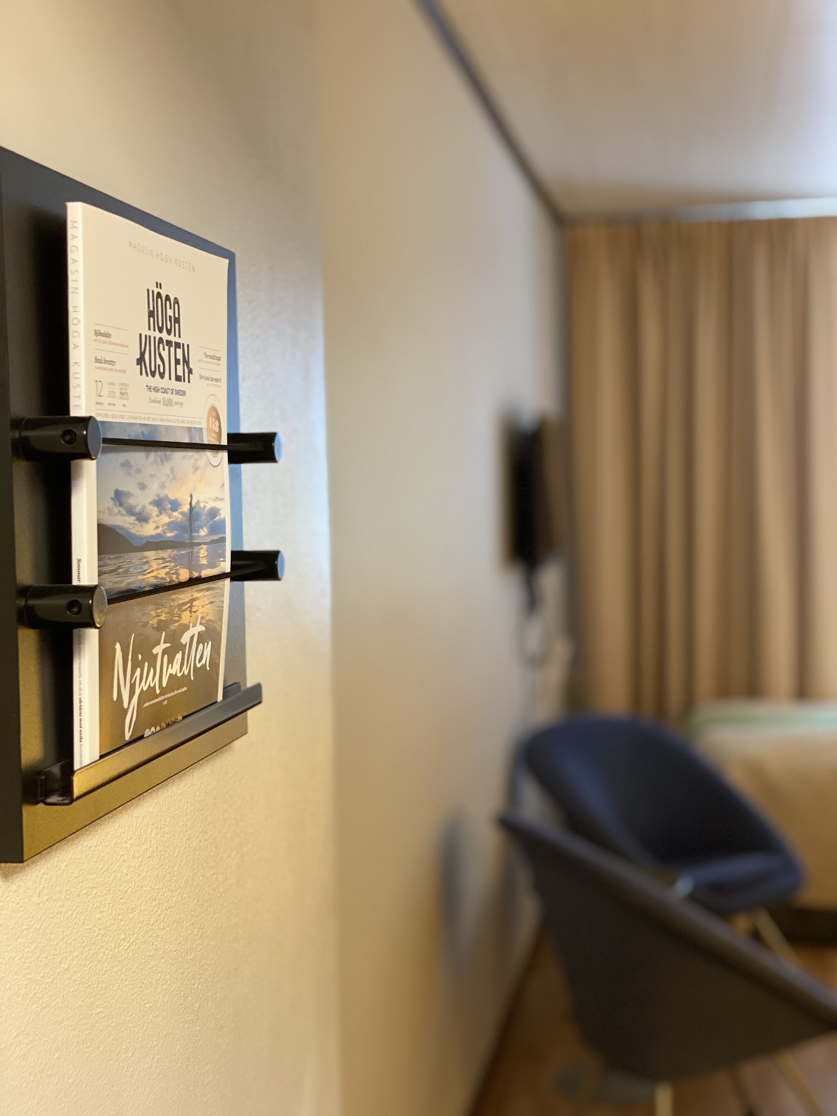 Quarto de hotel com estante na parede com livros, cama, cadeira e mesa de cabeceira.
