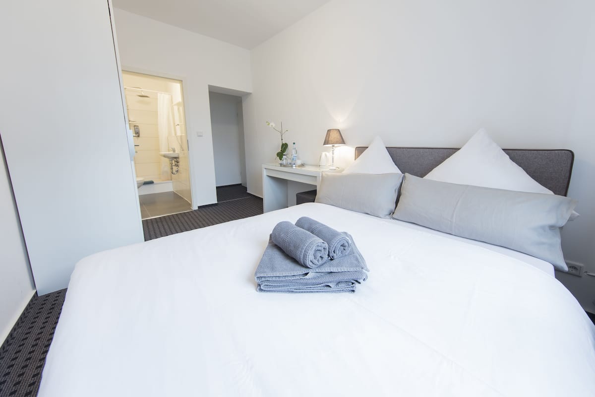 Quarto de hotel com cama bem arrumada, mesa de cabeceira com abajur, escrivaninha e vista para o banheiro.
