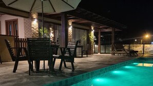 Uma área de piscina à noite, com piso ladrilhado e uma mesa com cadeiras sob um guarda-sol.