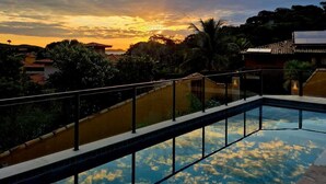 Piscina com barreira de vidro, cercada por vegetação e edifícios, sob um céu ao pôr do sol.