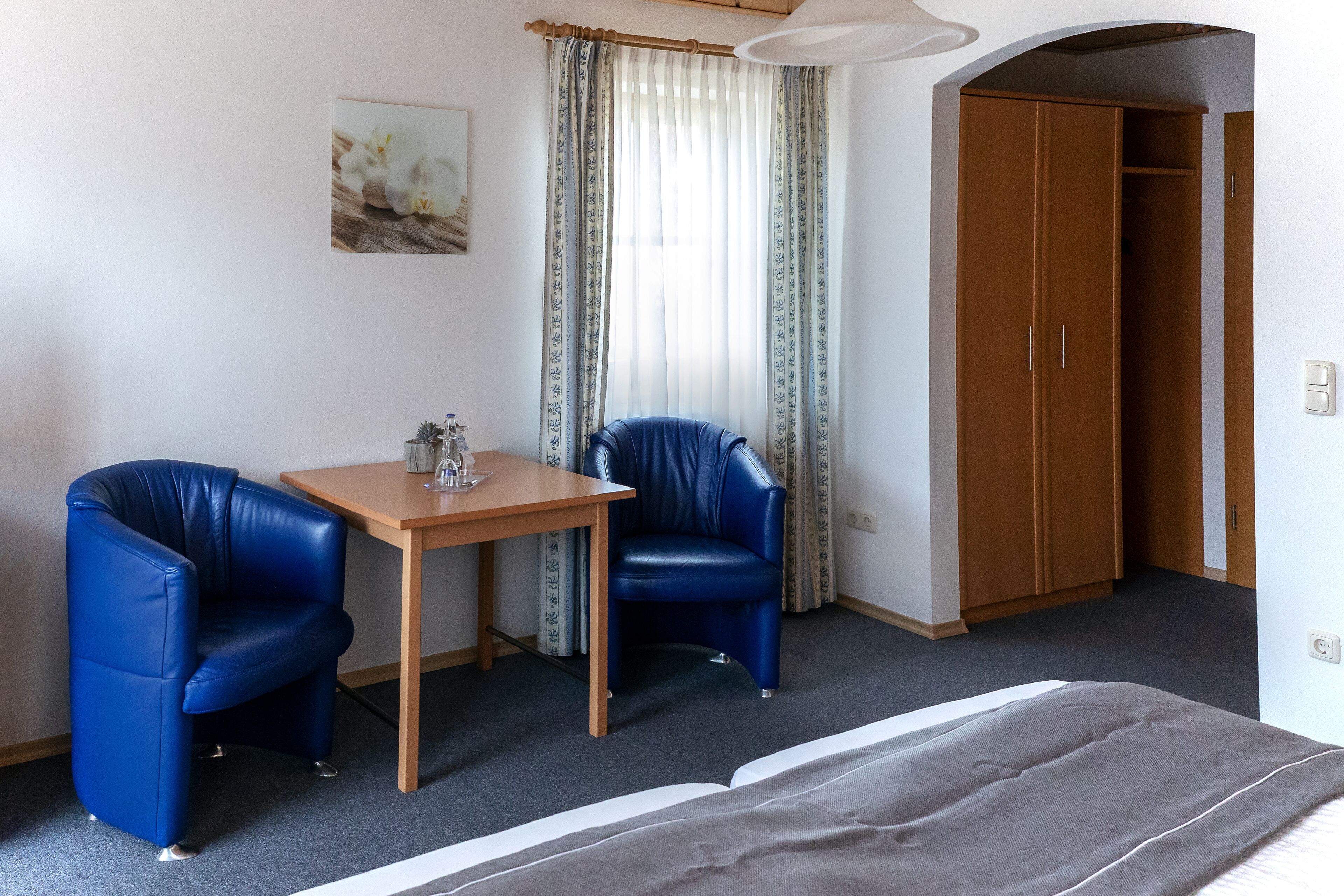 Quarto de hotel com uma cama, duas cadeiras azuis, uma mesa de madeira e um guarda-roupa.
