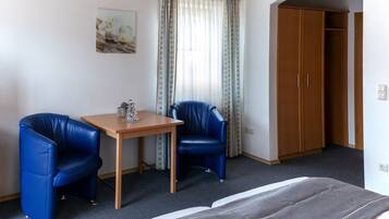 Quarto de hotel com uma cama, duas cadeiras azuis, uma mesa de madeira e um guarda-roupa.