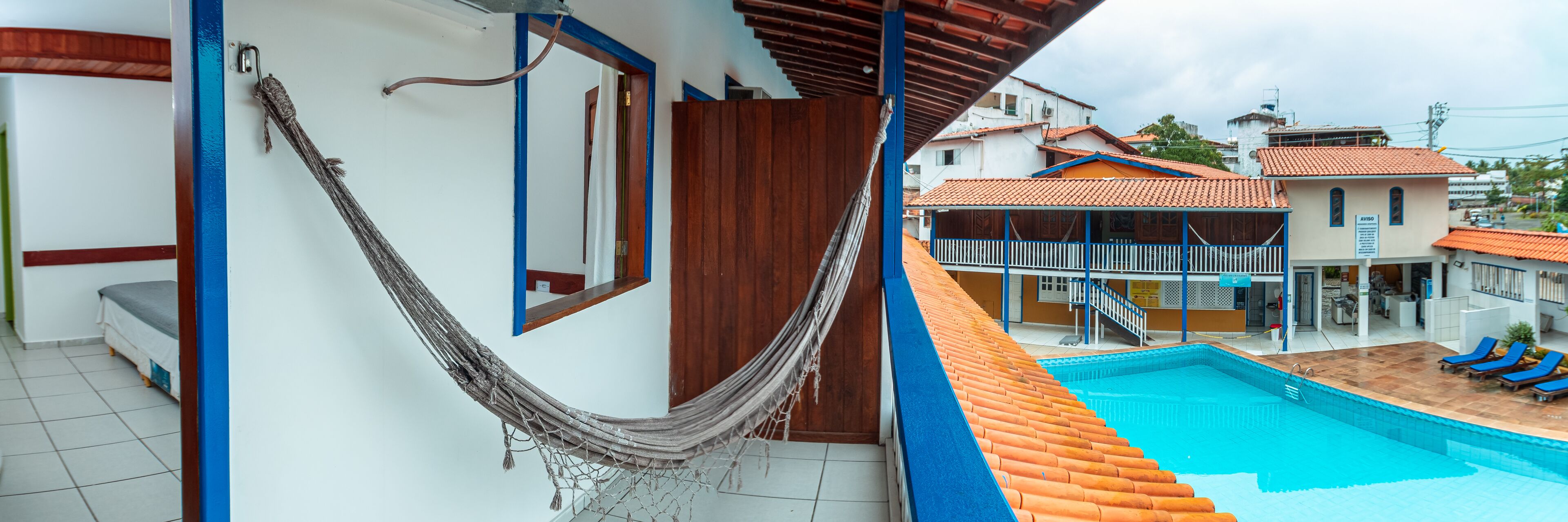 Uma varanda com rede, uma piscina com espreguiçadeiras azuis e vista para uma área residencial com casas.