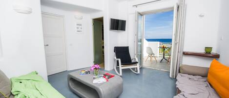 Uma sala de estar moderna com um sofá cinzento, um cobertor verde, uma cadeira branca e uma varanda com vista para o mar.