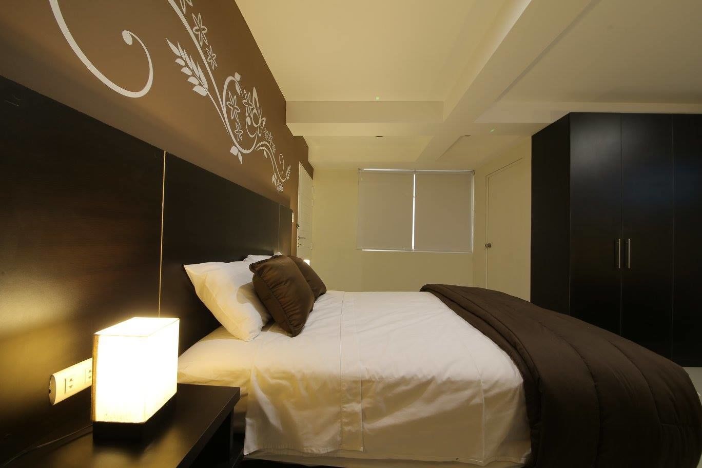 Quarto de hotel com cama, mesa de cabeceira com luminária, guarda-roupa e janela com persianas.