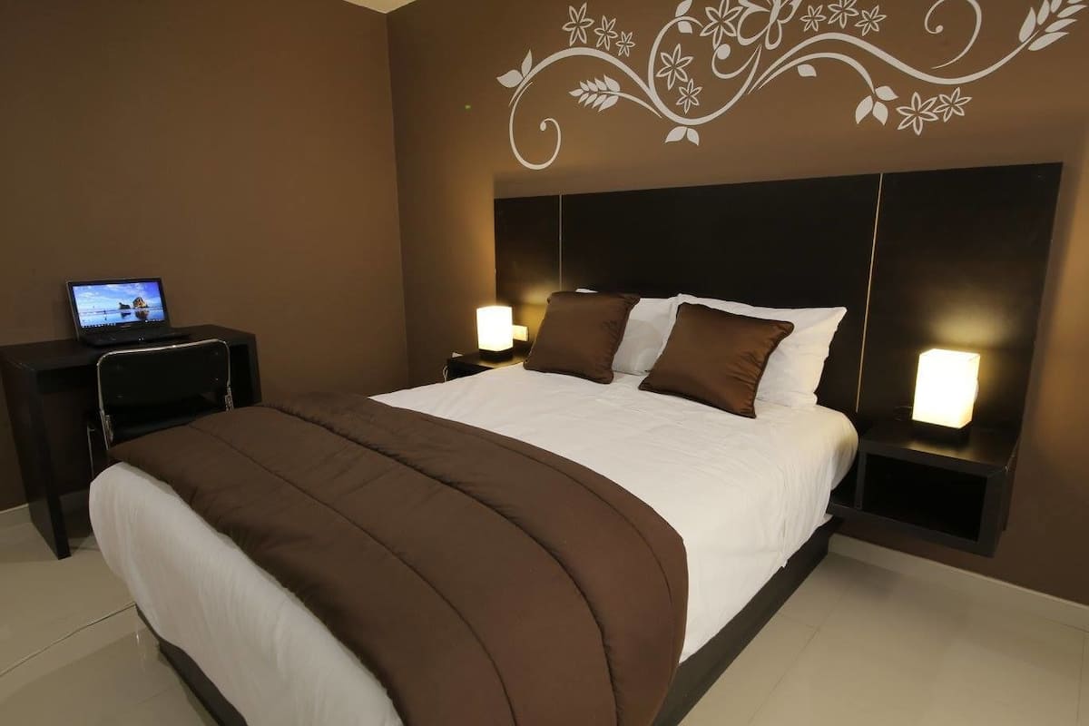 Quarto com uma cama grande, duas mesas de cabeceira com abajures, um laptop numa escrivaninha e um desenho decorativo na parede.