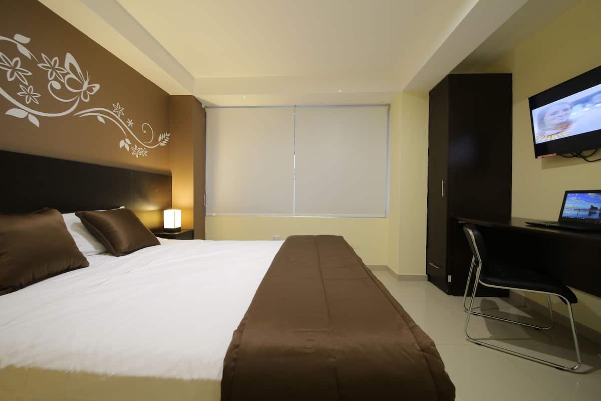 Quarto de hotel com uma cama grande, uma escrivaninha com cadeira, uma televisão e uma janela com persianas.