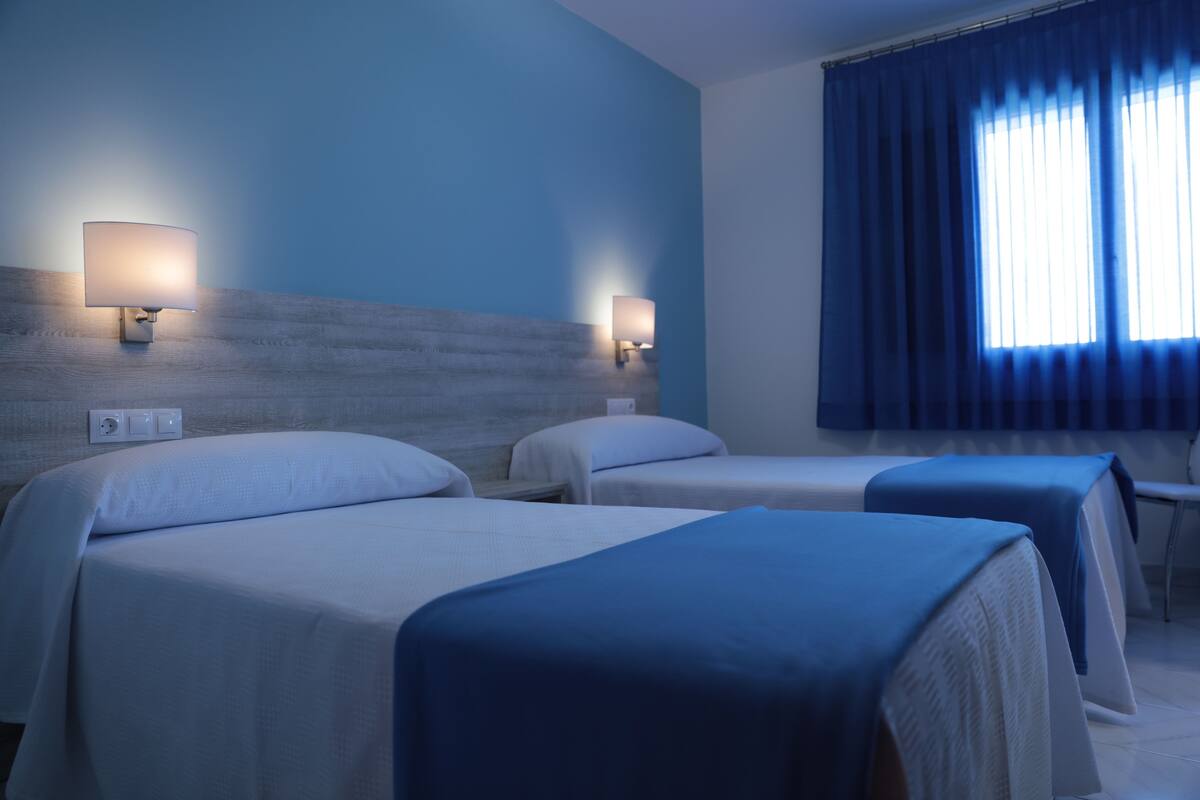 Quarto de hotel com duas camas, uma mesa de cabeceira, uma luminária e uma janela com cortinas.