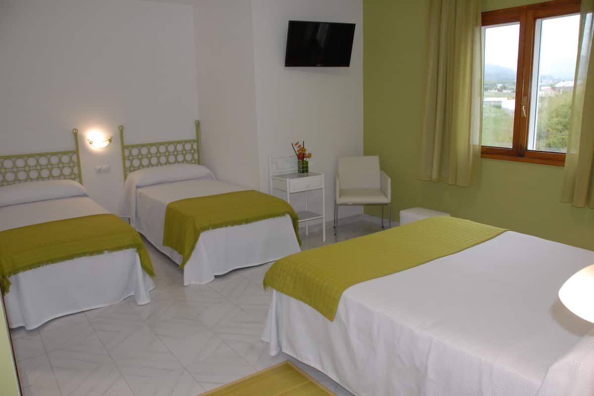 Quarto de hotel com três camas, uma pequena escrivaninha, uma cadeira e uma televisão na parede.