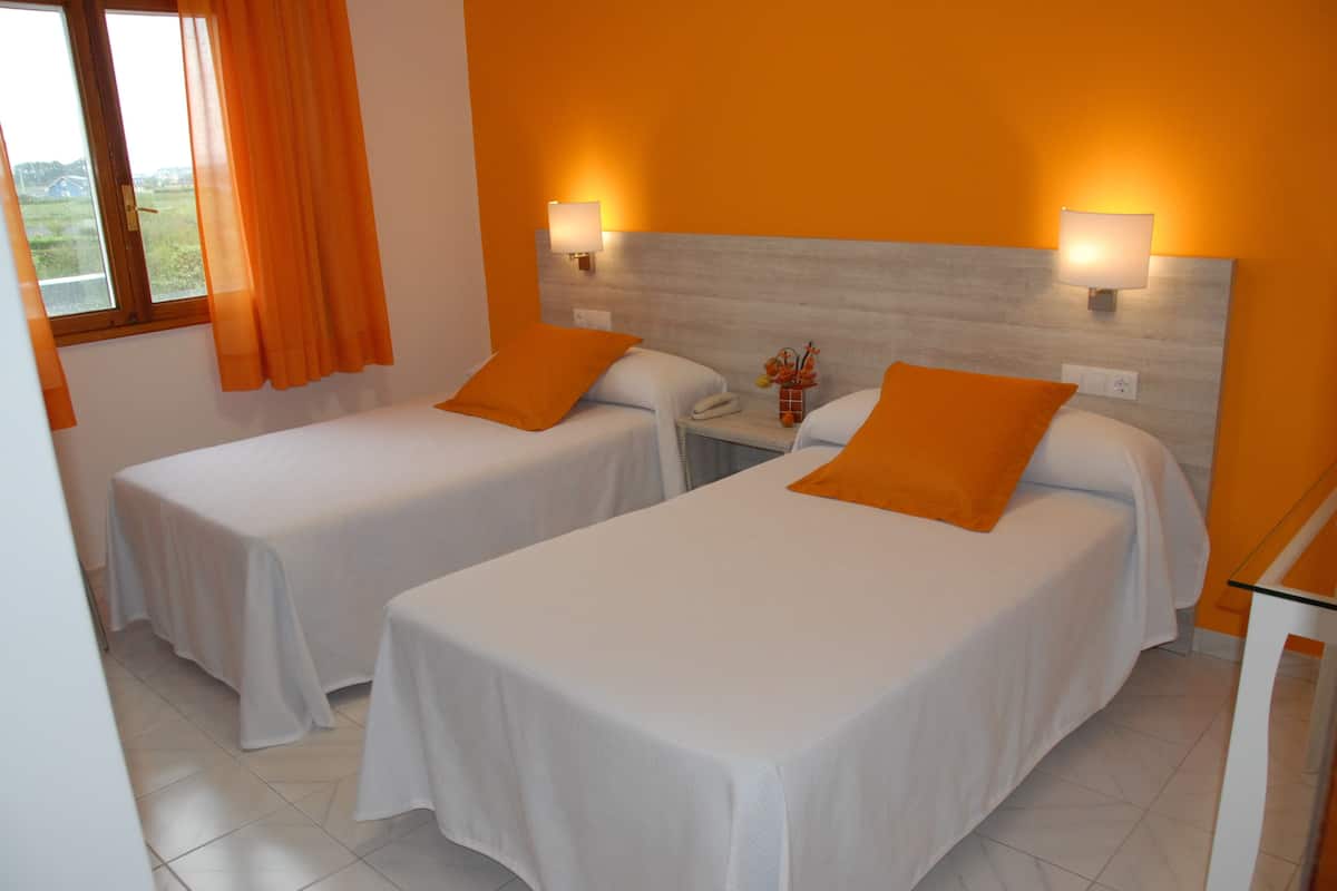 Quarto de hotel com duas camas de solteiro, cada uma com almofada laranja, roupa de cama branca e mesa de cabeceira com abajur.