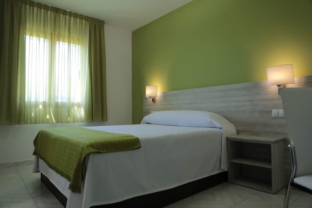 Quarto de hotel com cama, mesa de cabeceira, cadeira e janela com cortinas.