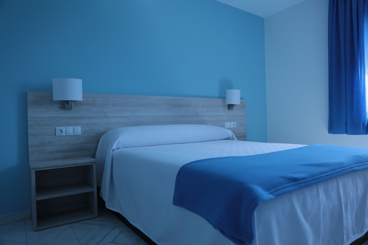 Quarto com cama, duas luminárias na parede, mesa de cabeceira e janela com cortinas azuis.