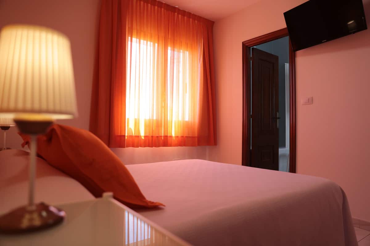 Quarto de hotel com cama, luminária, janela com cortinas e televisão fixada na parede.